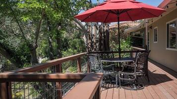 Um deck de madeira com uma mesa e cadeiras, um guarda-sol vermelho, e vista para árvores e uma casa.
