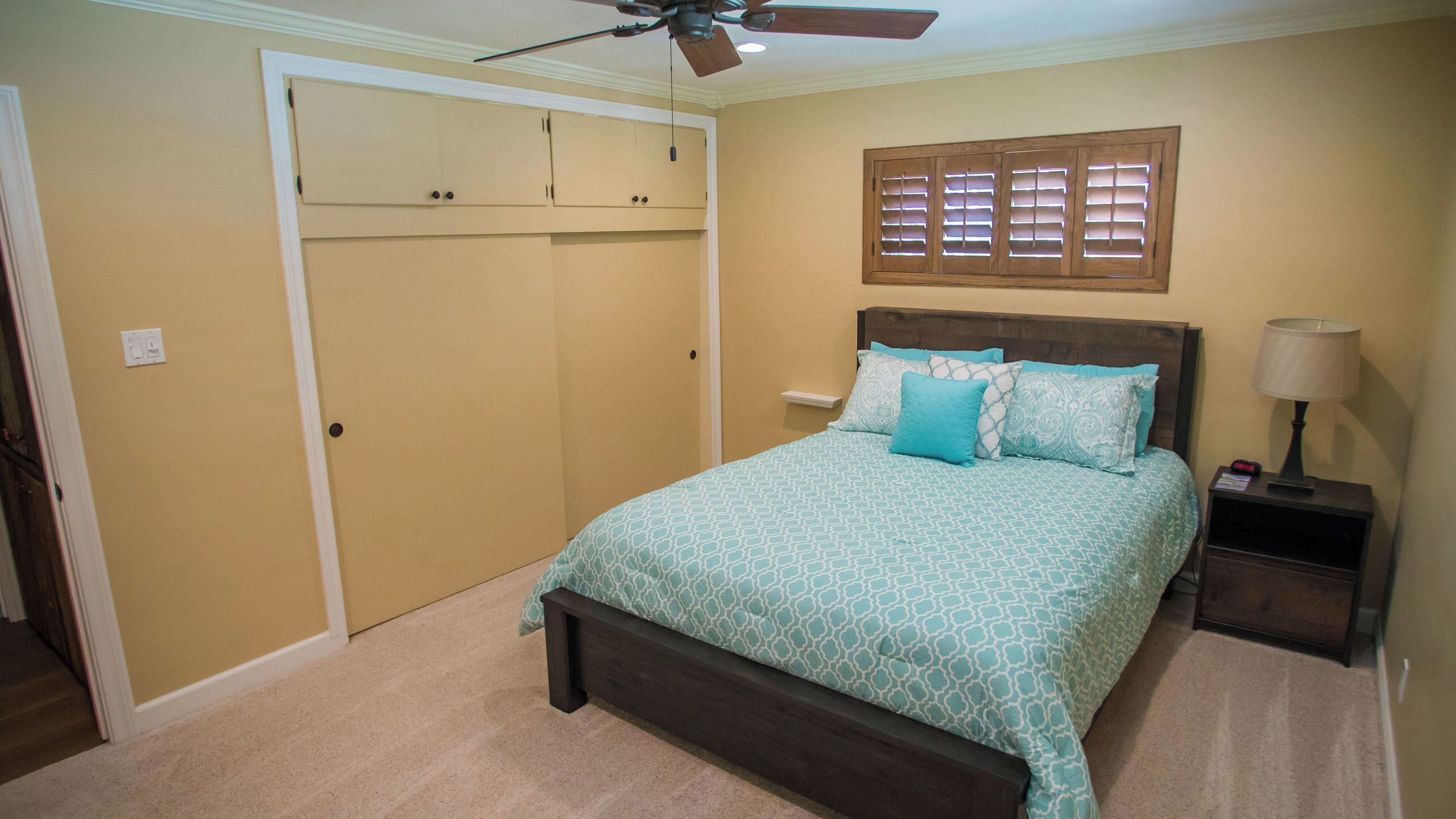 Quarto com cama, mesa de cabeceira, ventoinha no teto, janela com persianas e porta com armário.