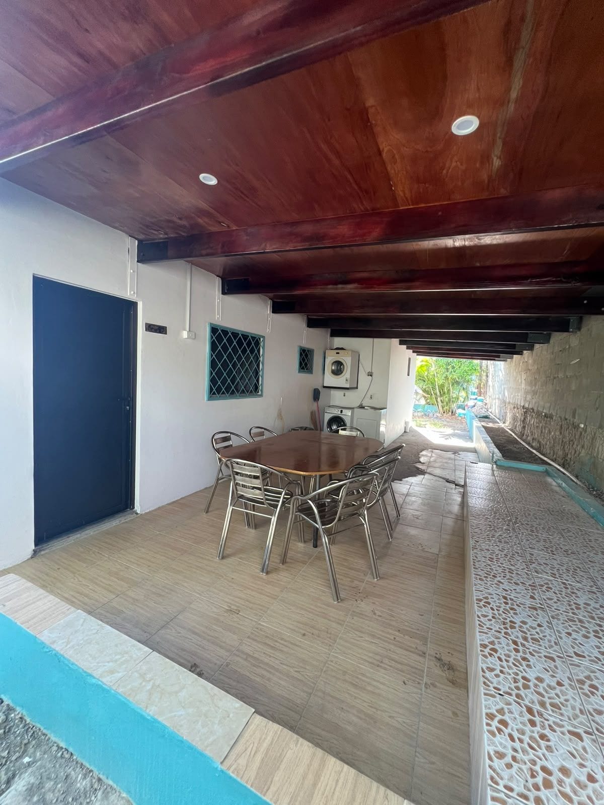 Uma área de refeições com mesa e cadeiras de madeira, uma porta azul e vista para o exterior.