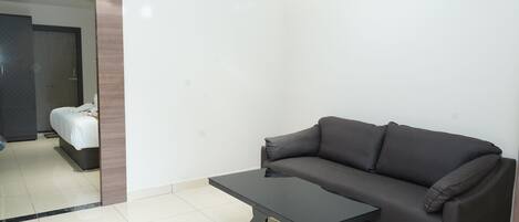 Uma sala de estar moderna com um sofá marrom escuro, uma pequena mesa de centro preta e uma cama com roupa de cama branca ao fundo.