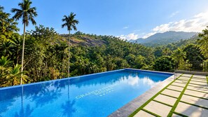 Piscina retangular com superfície de água azul cristalina, rodeada por vegetação exuberante e palmeiras, com montanhas ao fundo.