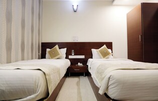 Quarto de hotel com duas camas, uma mesa de cabeceira, uma luminária na parede e um guarda-roupa de madeira.