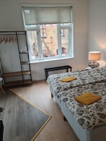 Quarto com cama, janela, mesa de cabeceira com abajur, piso de madeira e armário.