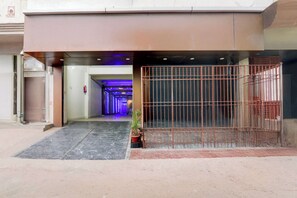 Entrada de um edifício com portão de metal, chão de azulejos e um corredor que leva a um interior bem iluminado.