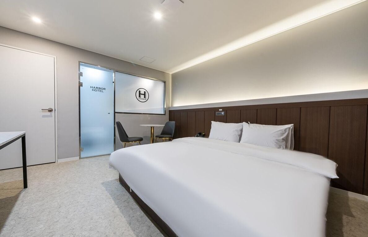 Quarto de hotel com uma cama grande, uma escrivaninha, uma cadeira e uma porta com a identificação 'Harbor Hotel'.