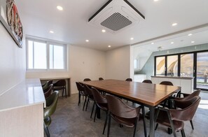 Uma sala de conferências com uma mesa comprida e cadeiras, teto branco com unidade de ventilação, e uma janela com vista para um edifício.