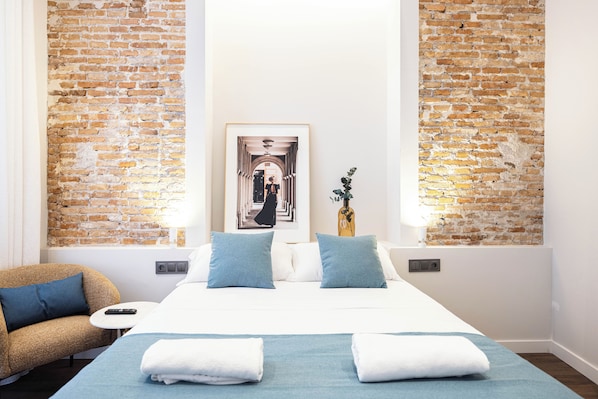Quarto de hotel com parede de tijolos, cama com roupa de cama azul, um sofá e um quadro pendurado na parede.