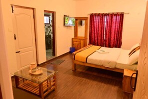 Quarto de hotel com uma cama, uma mesa de vidro, um armário de madeira, uma televisão e uma porta para o banheiro.
