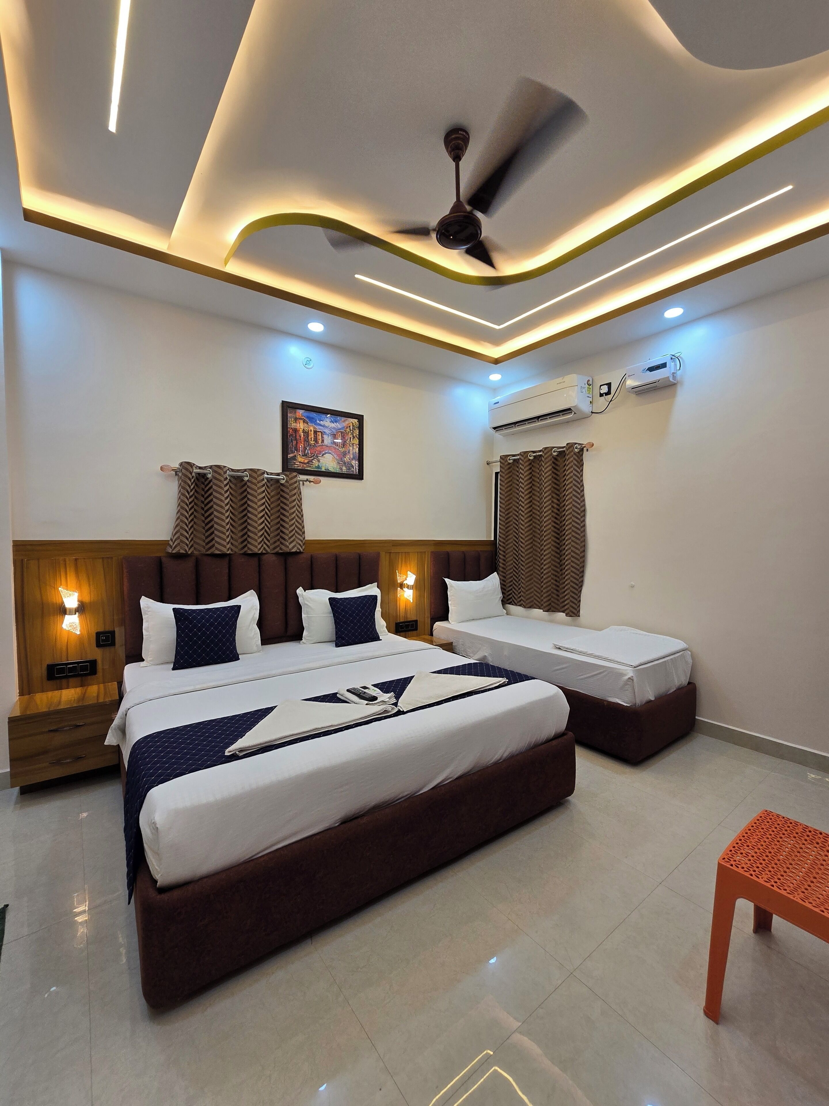 Quarto de hotel com uma cama grande, uma cama menor, um ventilador de teto, iluminação embutida, um aparelho de ar condicionado e um quadro na parede.