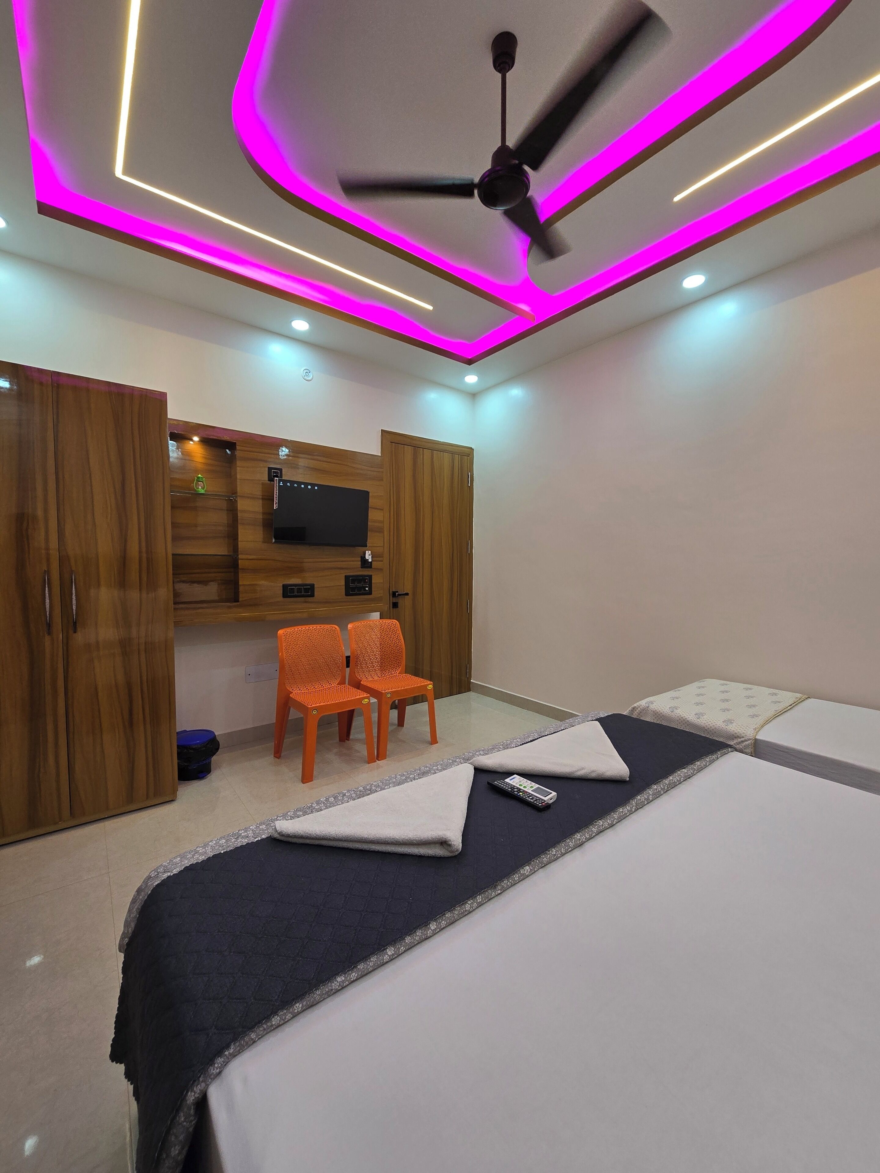 Quarto com cama, ventoinha no teto, televisão fixada em uma parede de madeira e duas cadeiras laranja.