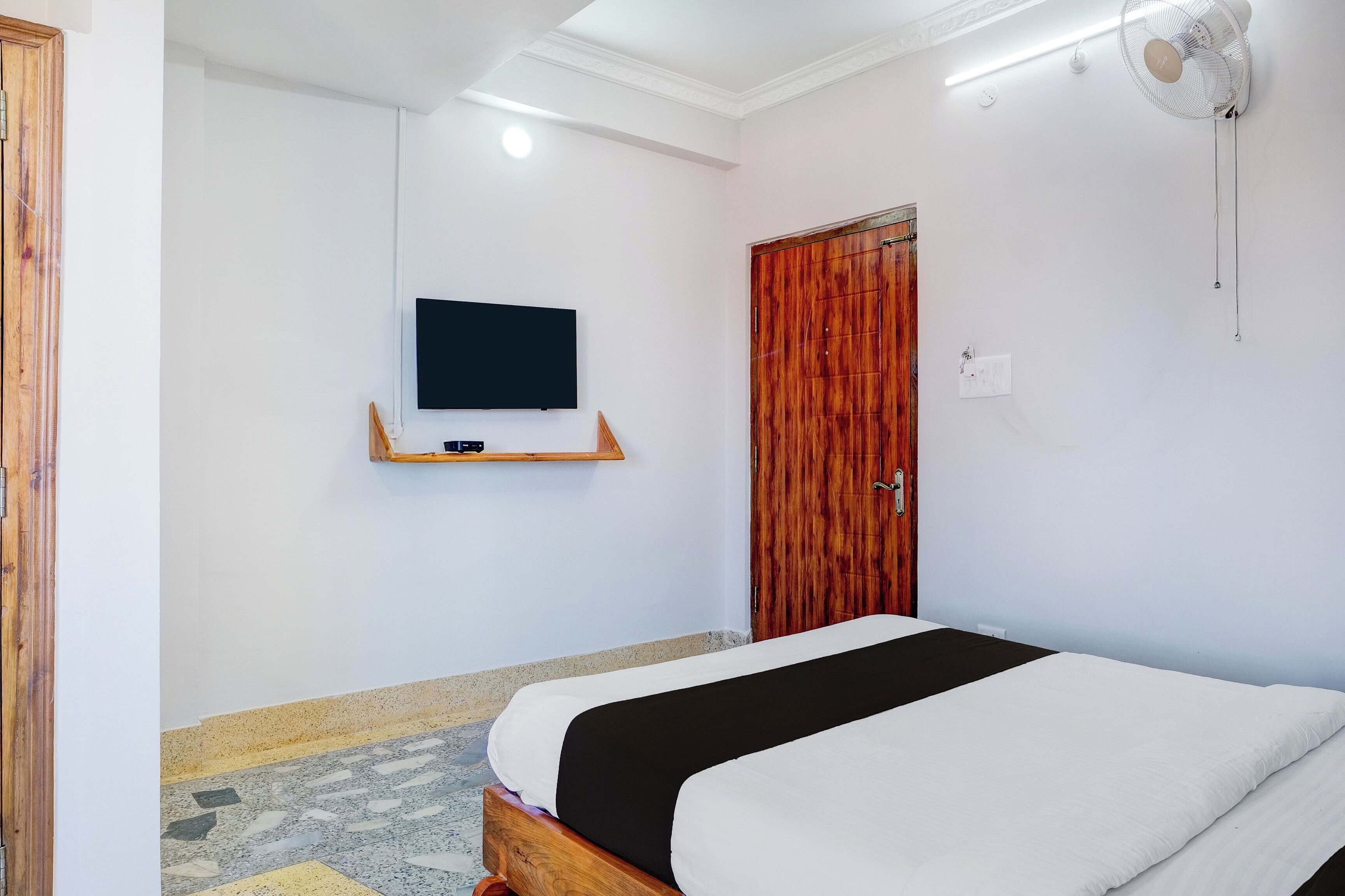 Quarto com cama, televisão na parede, porta de madeira e ventoinha no teto.