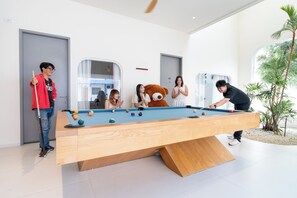 Uma mesa de bilhar em ambiente interno moderno, com uma grande decoração de urso de pelúcia, um ventilador de teto e um vaso de planta.
