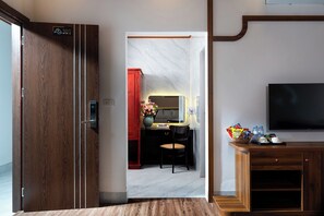Quarto de hotel com porta de madeira, escrivaninha com cadeira, televisão e um pequeno armário com diversos objetos.