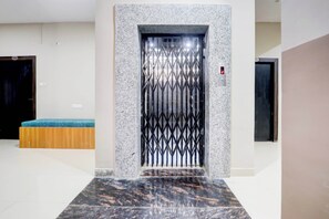 Um elevador moderno com portão de metal geométrico, piso de mármore e um banco azul ao fundo.