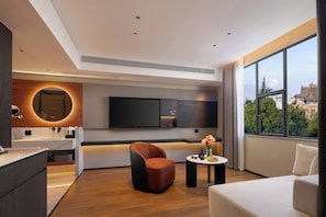 Quarto de hotel moderno com uma grande janela, uma TV de tela plana, um espelho redondo, um sofá e uma pequena mesa com um vaso de flores.
