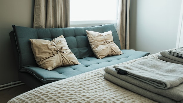 Quarto com sofá-cama, duas almofadas, um cobertor dobrado e uma janela com cortinas.
