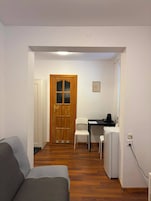 Quarto com porta de madeira, uma escrivaninha com cadeira, um pequeno frigorífico e um sofá.