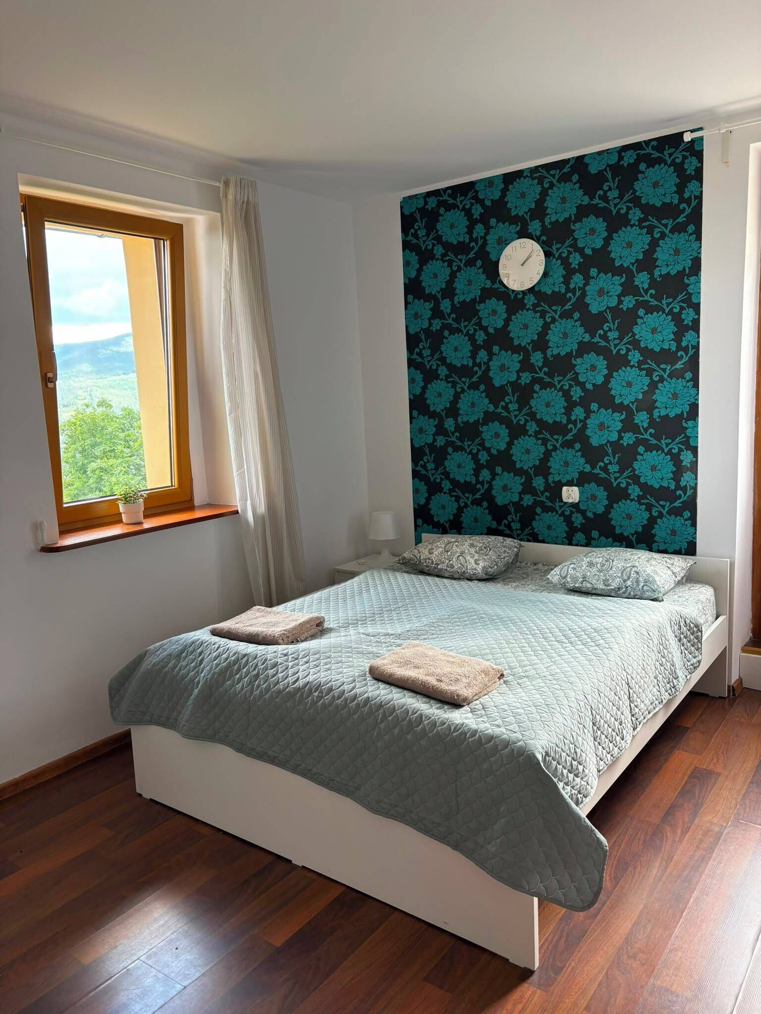 Quarto com cama, janela, relógio de parede e papel de parede floral.