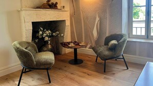 Um quarto com lareira, duas poltronas, uma mesa pequena com uma tigela e xícara, e uma janela com vista.