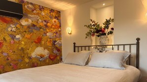 Um quarto com uma cama, um candeeiro fixado na parede e um vaso com flores.