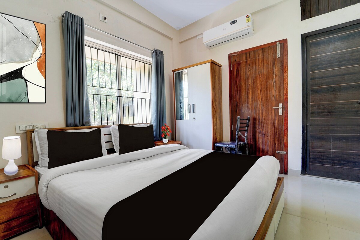 Quarto de hotel com cama, mesa de cabeceira, luminária, janela com cortinas, ar-condicionado e porta de madeira.