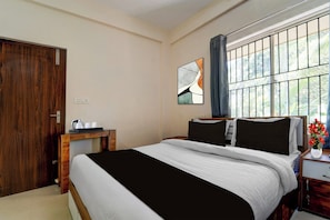 Quarto de hotel com cama, mesa de cabeceira com chaleira e xícaras, janela com cortinas e um quadro na parede.