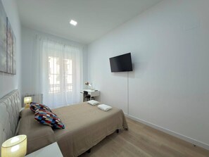 Quarto de hotel com cama, escrivaninha, cadeira, televisão fixada na parede e janela com cortinas.