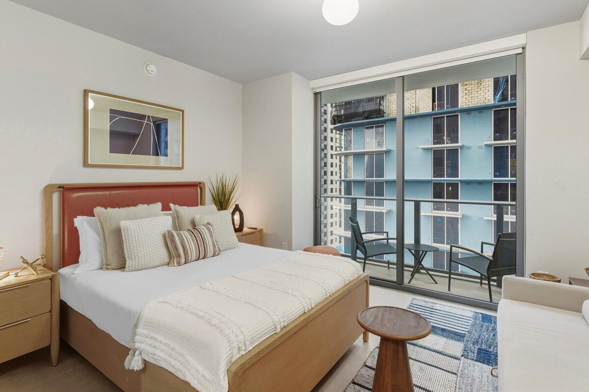 Quarto moderno com cama grande, mesa de cabeceira, cadeira, pequena mesa redonda e varanda com vista para um edifício.