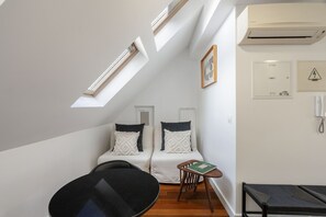 Quarto moderno e compacto com cama em mezanino, uma pequena mesa e uma cadeira. Possui claraboias, ar condicionado e telefone fixo na parede.