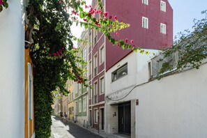 Rua estreita com edifícios de ambos os lados, alguns com varandas e outros com entradas diretas.
