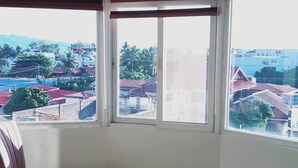 Quarto de hotel com uma cama, duas cadeiras, uma mesinha e grandes janelas com vista para a cidade.