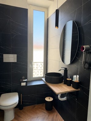 Casa de banho moderna com parede de mármore preto, lavatório redondo, sanita e janela com vista.