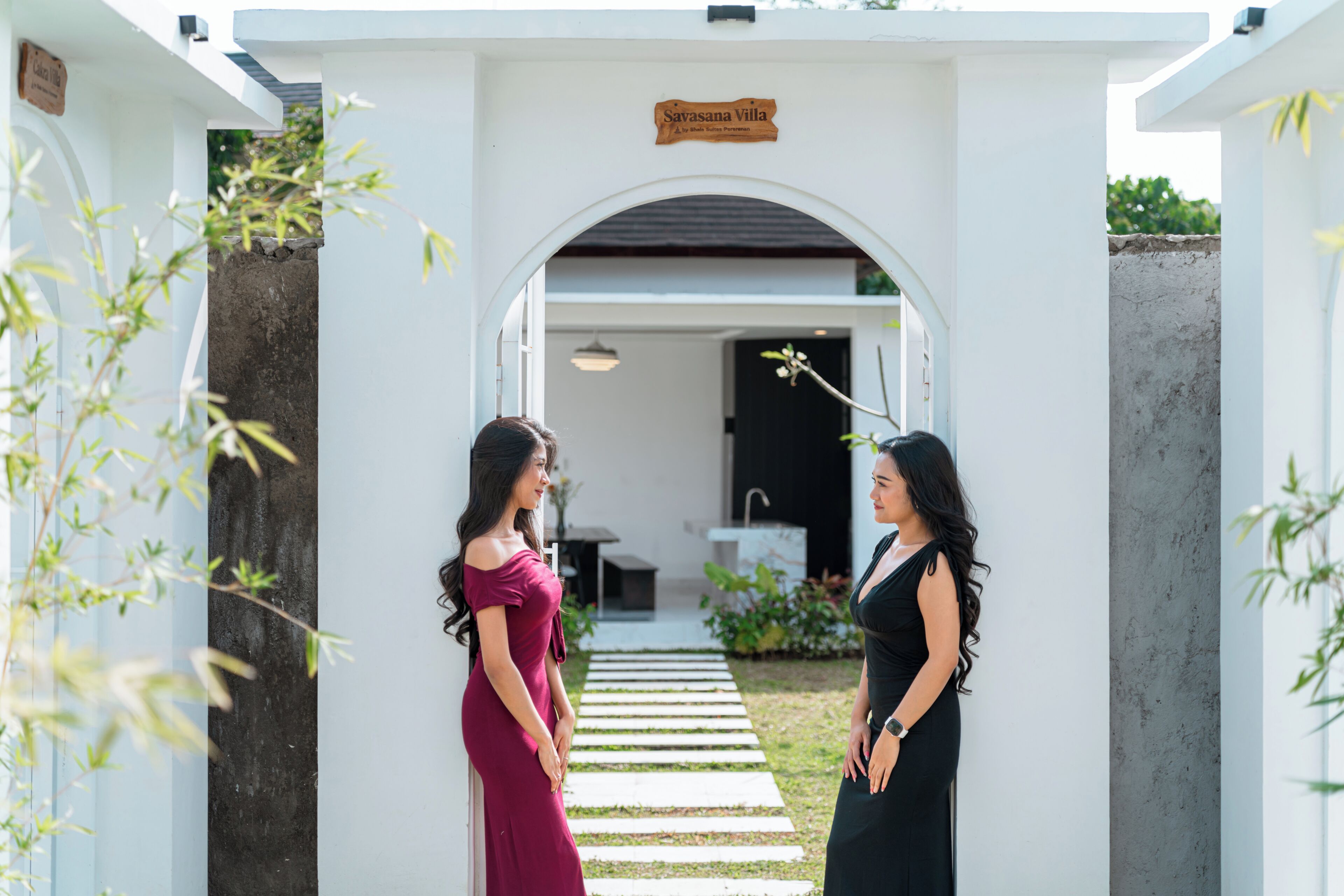Um arco branco com uma placa indicando 'Savasana Villa' que dá acesso a um espaço interior moderno, com área de refeições e uma escadaria.