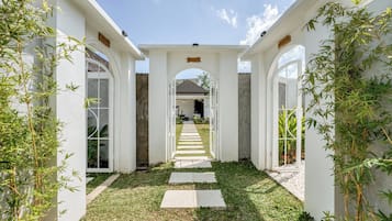 Um caminho leva a um edifício com paredes brancas e portas de vidro, rodeado por vegetação.