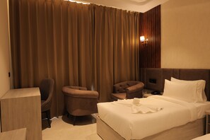 Quarto de hotel com cama, duas poltronas, escrivaninha e uma janela grande com cortinas.