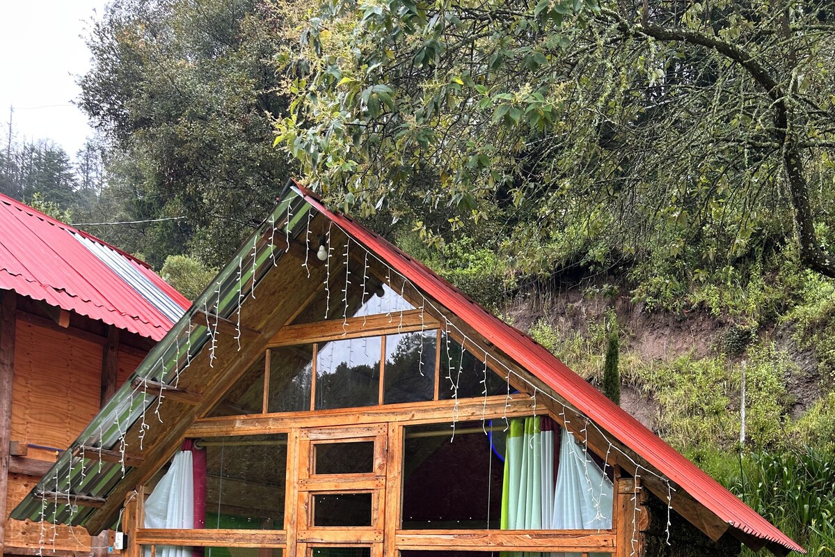 Uma cabana de madeira com telhado vermelho, janelas amplas e uma escadaria de madeira que leva à entrada.