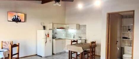 Quarto com teto de madeira, cama com colcha acolchoada, escrivaninha com cadeira e cozinha ao fundo.
