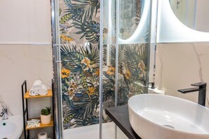 Uma casa de banho com uma grande pia branca, cabine de chuveiro em vidro com papel de parede com folhas e flores tropicais, um porta-toalhas com toalhas e um pequeno armário com produtos de higiene pessoal.