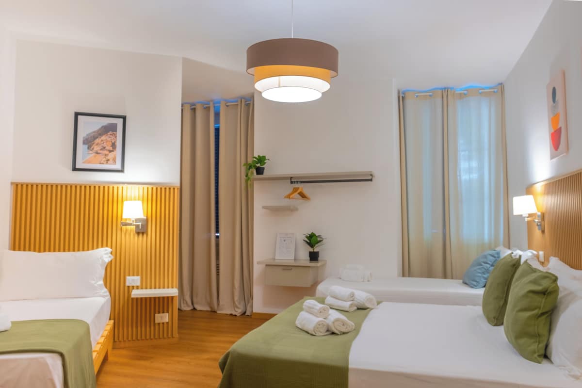 Quarto de hotel com duas camas, mesa de cabeceira, luminária, quadro pendurado na parede e janela com cortinas.