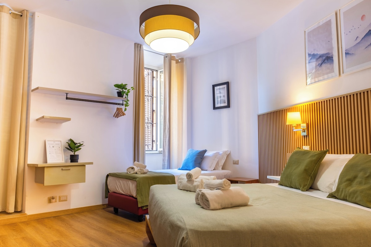 Quarto de hotel com uma cama, duas mesas de cabeceira, uma janela com cortinas, uma prateleira fixa com uma planta e um quadro emoldurado.