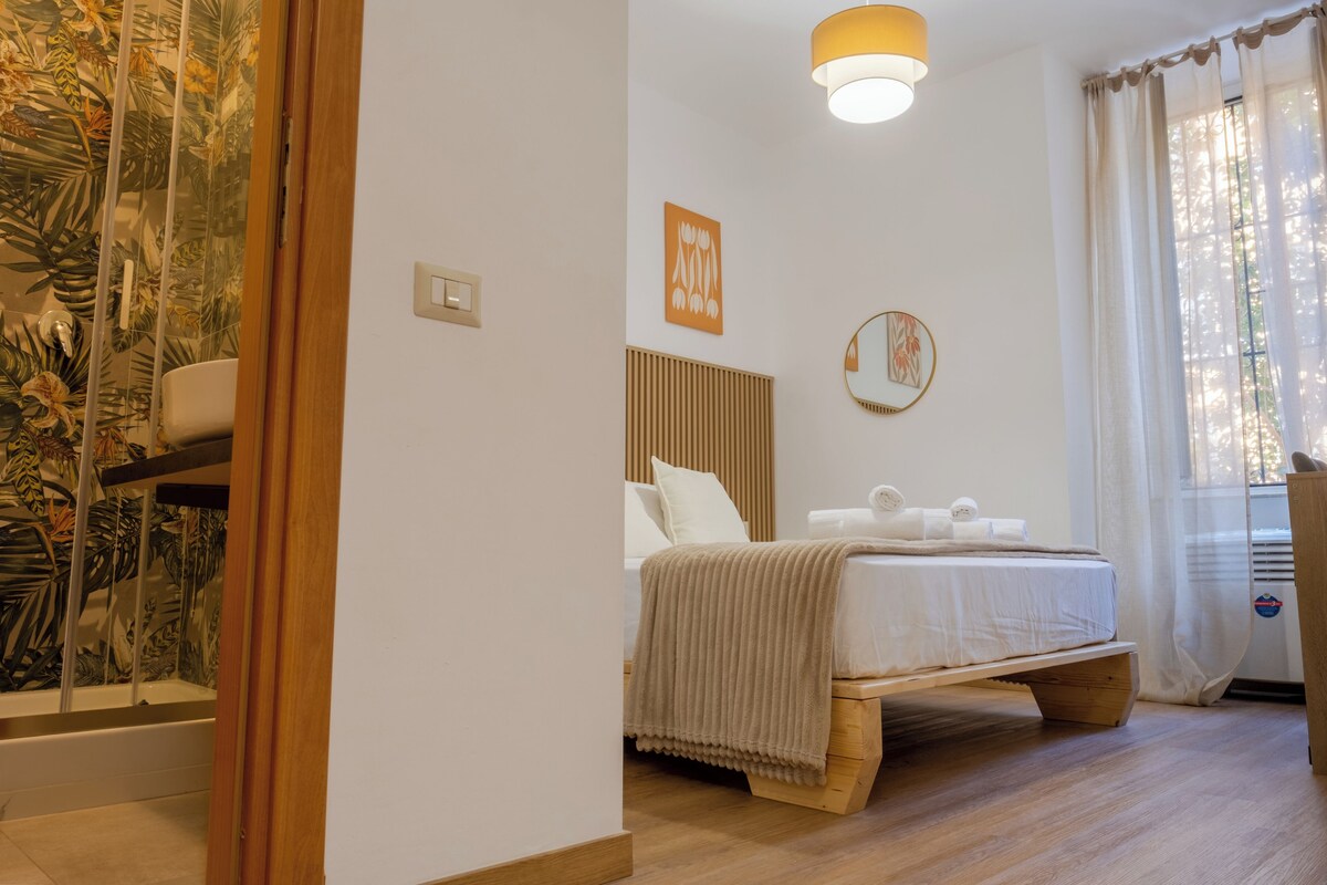 Quarto com cama, mesa de cabeceira, luminária na parede e janela com cortinas.