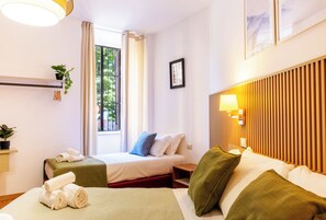 Quarto de hotel com duas camas, janela com cortinas, mesa de cabeceira com abajur e prateleira na parede com uma planta.