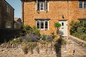 A stone building with a traditional design, featuring a central window with shutters, a door, and a small garden area with various plants.