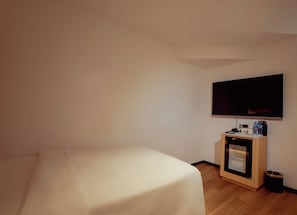 Quarto de hotel com uma cama, televisão fixada na parede, pequeno armário de madeira com um cesto de lixo e um dispensador de água.