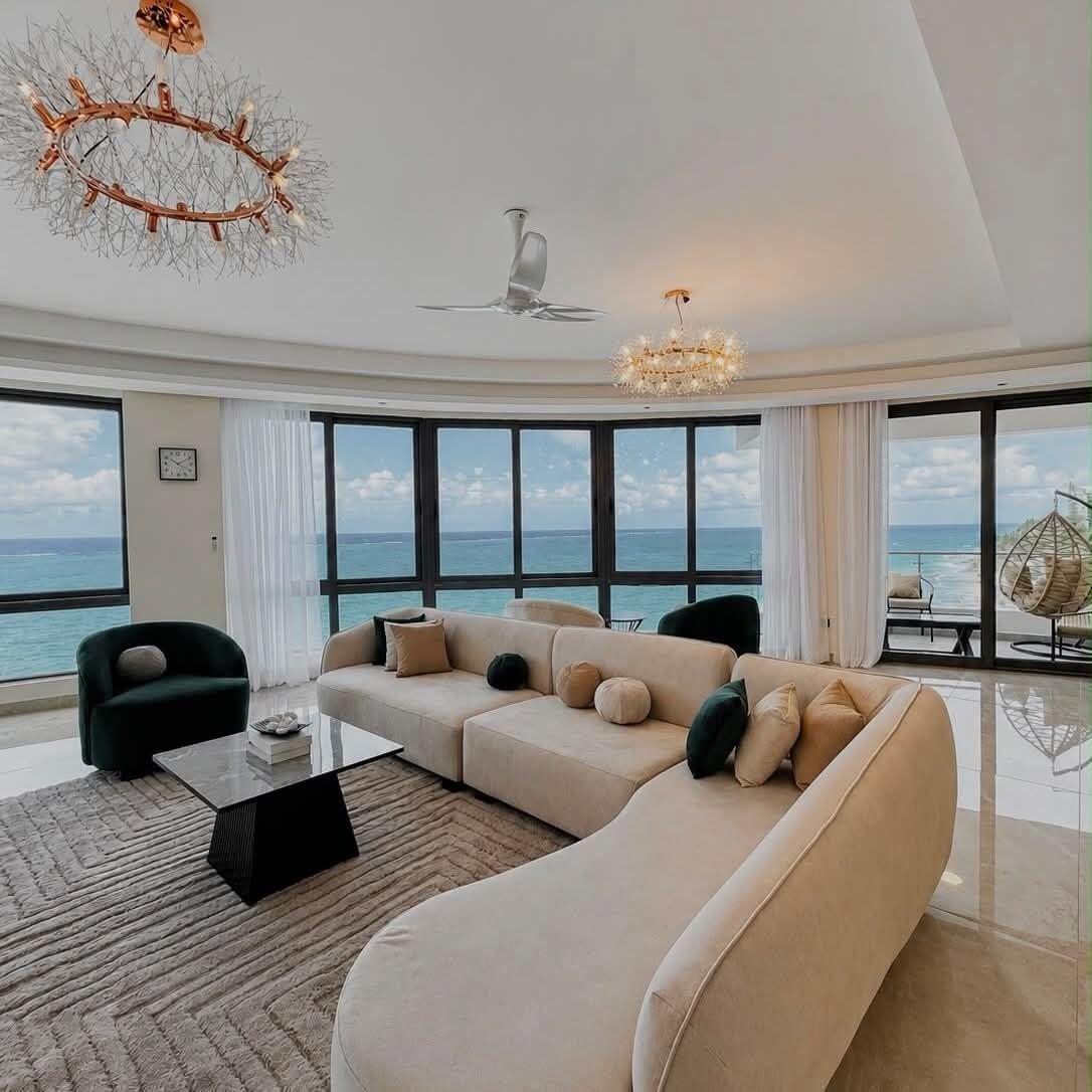 Uma sala de estar moderna com um sofá grande e curvo, uma mesa de centro e vista para o oceano através de amplas janelas.
