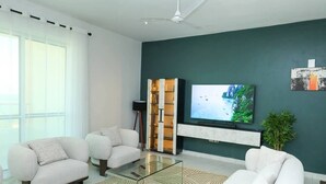 Uma sala de estar moderna com televisão de tela plana, sofás brancos, uma mesa de centro de vidro e um ventilador de teto.