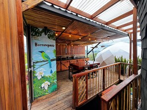 Uma área de refeições ao ar livre com um deck de madeira, um mural com aves e folhagens tropicais, e vista para a paisagem.