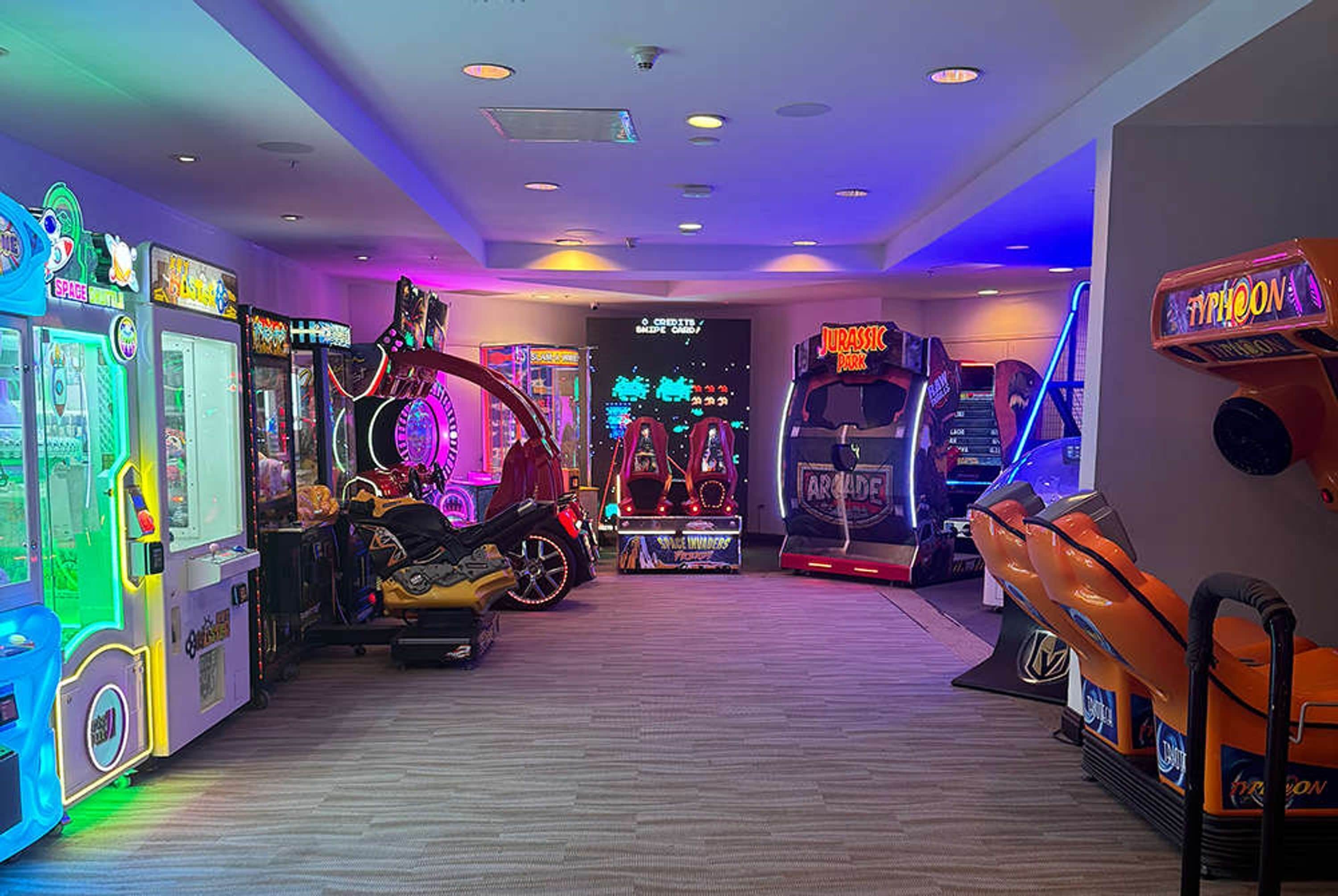 Um salão de jogos indoor com diversas máquinas de entretenimento, incluindo uma atração temática de 'Jurassic Park'.