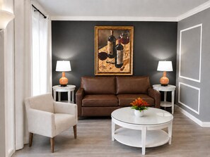 Uma sala de estar moderna com um sofá de couro marrom, duas mesas laterais com abajures, uma mesa de centro redonda branca com um vaso de flores e uma obra de arte emoldurada na parede.