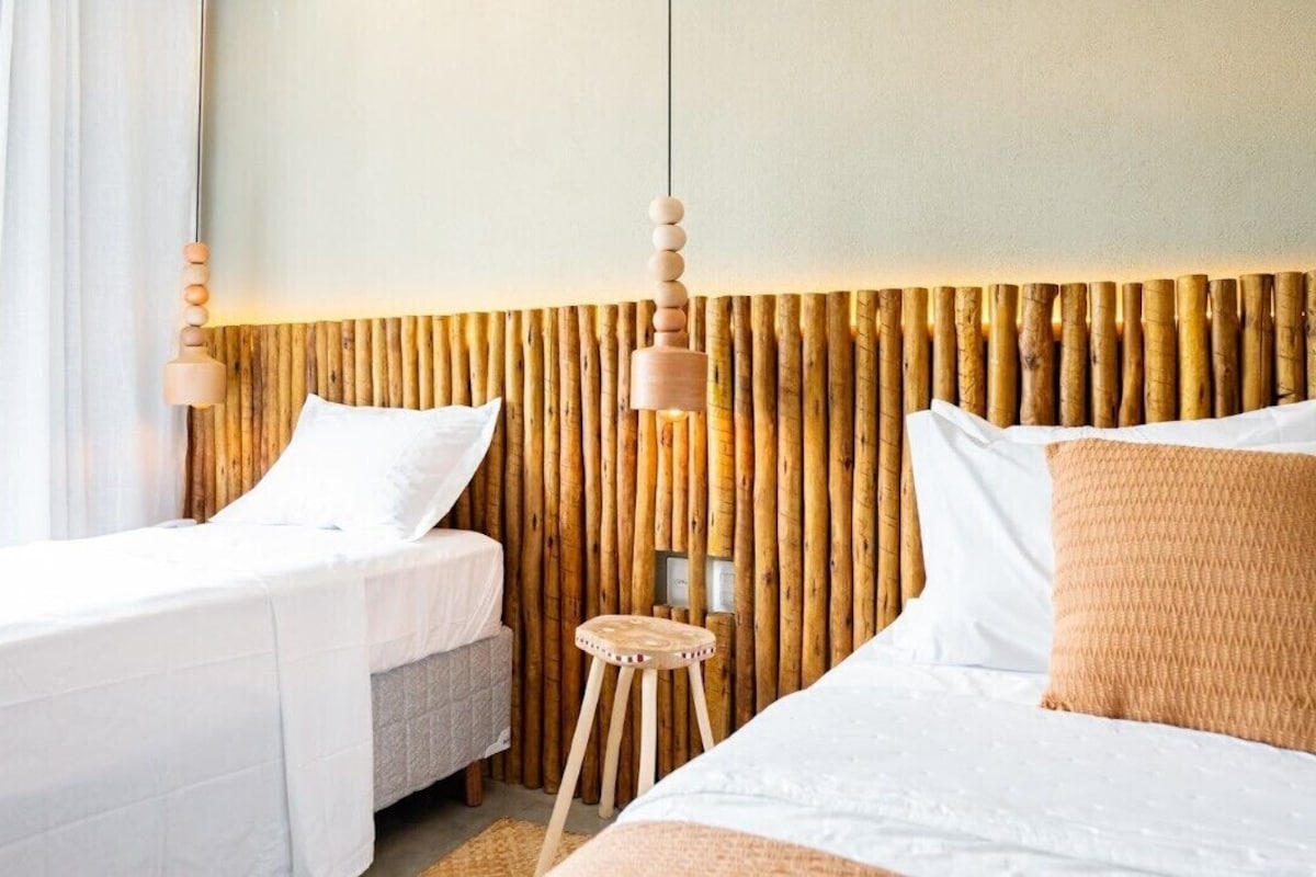 Quarto de hotel com duas camas, cabeceira de madeira, um banquinho pequeno e luminária pendente.